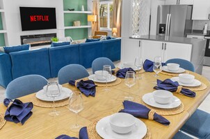 Uma sala de jantar com mesa posta para seis pessoas, rodeada por cadeiras azuis, uma cozinha moderna ao fundo e uma TV exibindo o logotipo da Netflix.