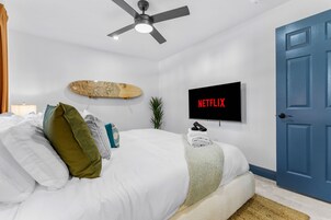 Quarto com cama, almofadas, televisão na parede, ventoinha de teto e uma porta.