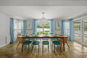 Uma sala de jantar com uma mesa posta para seis pessoas, rodeada de cadeiras, e vista para uma piscina e áreas verdes lá fora.