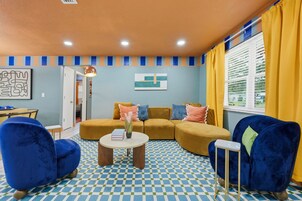 Uma sala de estar com piso xadrez, um sofá, duas poltronas azuis, uma mesinha de centro e uma janela com cortinas amarelas.