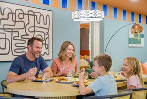Uma família jantando em uma mesa redonda, em um ambiente interno moderno, com uma obra de arte geométrica na parede e um lustre pendurado.