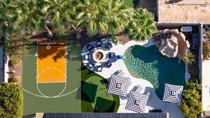 Vista aérea de um resort com quadra de basquete, área de estar externa, piscina e palmeiras.