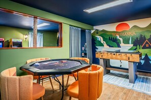 Um quarto com mesa de bilhar, quatro cadeiras laranjas, mesa de futebol de mesa e um mural com o texto 'TOBYHANNA', além de uma janela com cortinas.