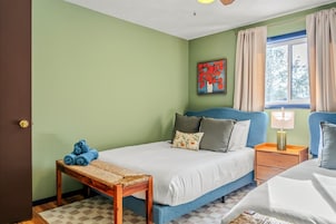 Um quarto com cama, mesa de cabeceira, um banco, janela com cortinas e um quadro na parede.