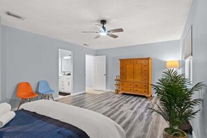 Quarto com cama, duas cadeiras, cômoda de madeira, ventilador de teto e planta em vaso.