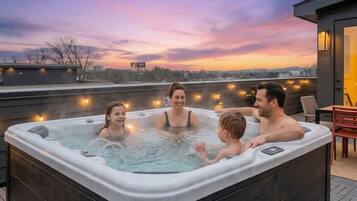 Uma família aproveitando uma banheira de hidromassagem em um terraço com vista para a cidade ao pôr do sol.