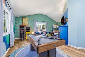 Uma mesa de bilhar em um quarto com parede estampada em verde, um sofá e uma televisão exibindo o logotipo da Netflix.