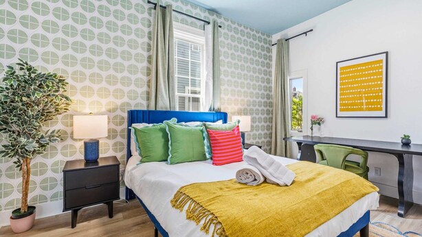 A hotel room with a bed, bedside table, lamp, potted plant, desk, chair, and a framed picture on the wall.