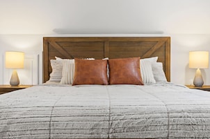 Uma cama com cabeceira de madeira, duas mesas de cabeceira com abajures e várias almofadas.