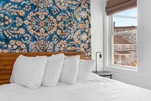 Quarto com cama grande, roupa de cama branca, cabeceira de madeira, tapeçaria de parede estampada e janela com vista para prédios.