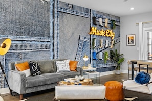 Uma sala de estar moderna com um sofá cinzento, uma mesa de centro com livros e um globo, uma parede com textura jeans e um letreiro com a inscrição 'Music City', e uma área de jantar com mesa e cadeiras.