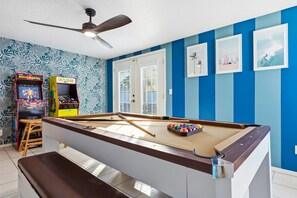 Uma mesa de bilhar, um ventilador de teto, jogos de fliperama e quadros em uma parede azul.