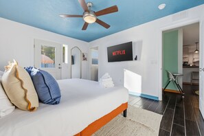 Quarto com cama, ventilador de teto, televisão fixada na parede e porta de acesso a um banheiro.