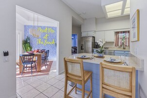 Cozinha moderna com ilha central, armários brancos e área de refeições com parede azul e uma placa com a inscrição 'Meet Me at The Beach'.