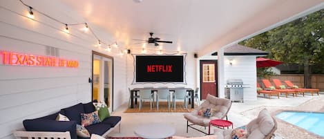 Um pátio externo com piscina, área de estar e uma televisão exibindo 'NETFLIX'.