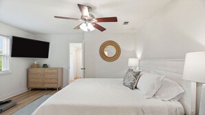 Quarto com cama, cômoda, televisão, ventoinha no teto, espelho e luminária de cabeceira.