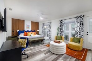 Quarto moderno com cama, escrivaninha, cadeira, um banquinho redondo e janela com cortinas estampadas.
