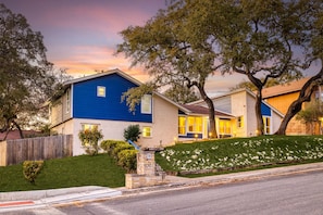 Uma casa residencial de dois andares com fachada azul, um jardim frontal e vista para a rua.