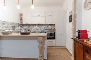Uma cozinha com armários brancos, uma ilha central com cúpula de vidro, uma cafeteira vermelha e um revestimento de azulejos.