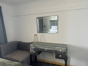 Um quarto com um sofá cinzento, um penteou espelhado com gavetas e uma pequena mesa com abajur.