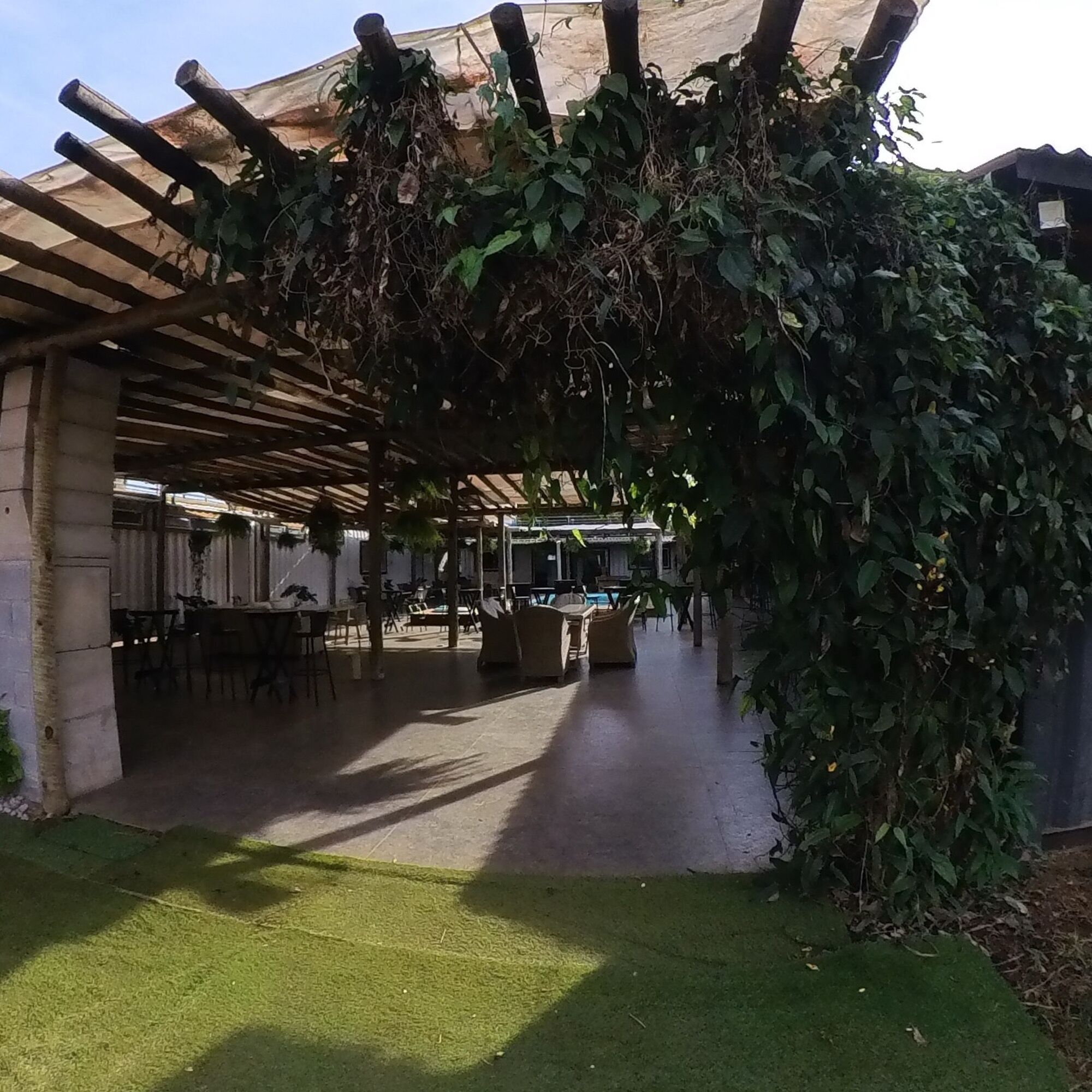 Área externa coberta com mesas e cadeiras, piscina e uma pérgula com plantas trepadoras.