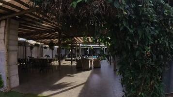 Área externa coberta com mesas e cadeiras, piscina e uma pérgula com plantas trepadoras.