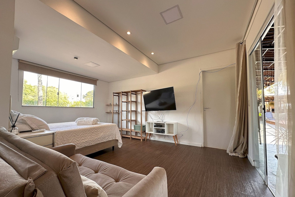 Quarto de hotel moderno com uma cama grande, um sofá, uma TV na parede e uma janela com vista para o verde.