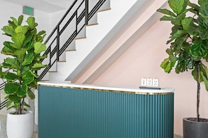 Bancada de recepção com um visor de data, ladeada por plantas em vasos, em um edifício moderno com uma escadaria.