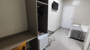 Quarto compacto com uma cama, escrivaninha, cadeira, guarda-roupa e televisão na parede.