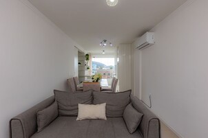 Uma sala de estar moderna com um sofá cinzento, uma área de jantar com mesa e cadeiras, e vista para o exterior.