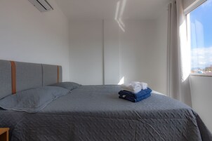 Cama bem arrumada com edredom e almofadas cinzas, janela com cortinas, e vista para prédios lá fora.