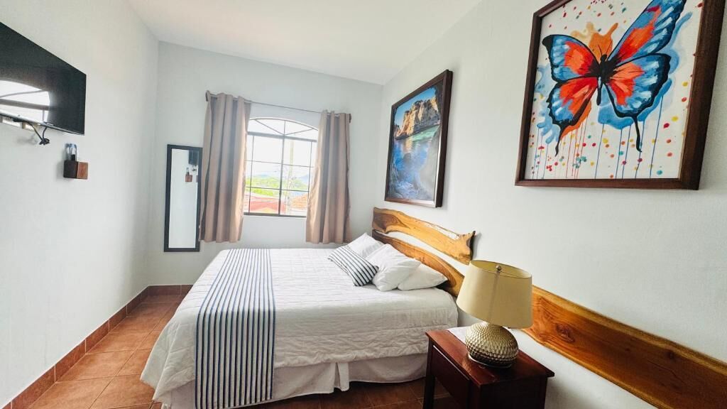 Quarto com cama, mesa de cabeceira com abajur, televisão presa à parede e janela com cortinas.
