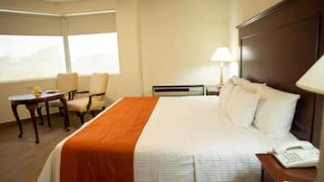 Quarto de hotel com cama, escrivaninha, cadeira, luminária e janela.