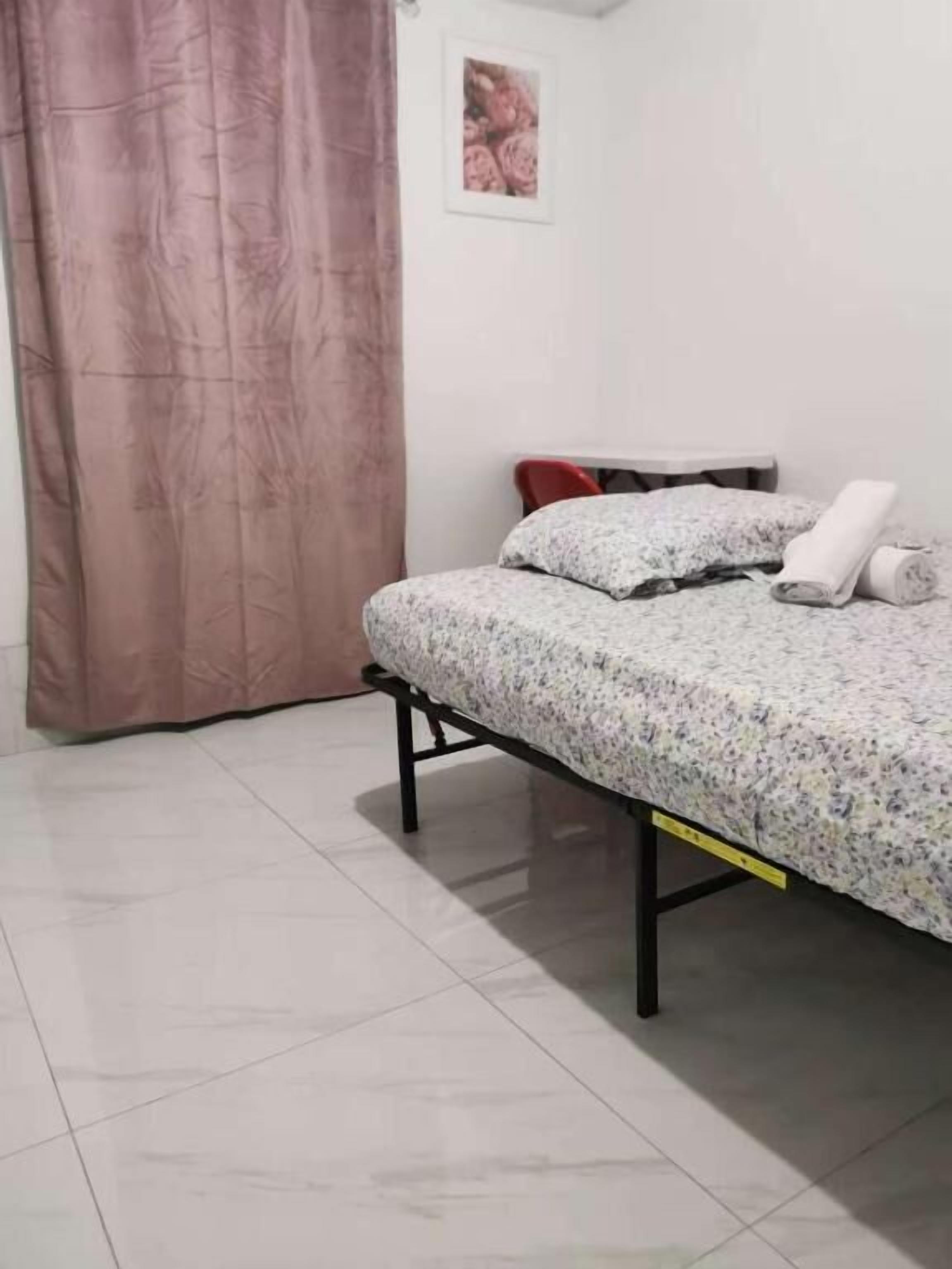 Quarto com cama, travesseiro, toalha enrolada, cortina e uma pequena fotografia na parede.