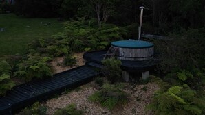 A circular hot tub surrounded by a wooden deck and ferns, with a chimney-like structure nearby.