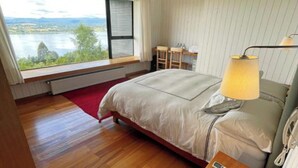 A bedroom with a bed, a desk, a chair, a lamp, and a view of a lake.