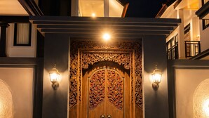 Uma entrada escura com porta de madeira adornada com esculturas detalhadas e duas luzes fixadas na parede.