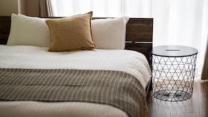 Uma cama bem arrumada com colcha listrada e duas almofadas, uma mesinha de cabeceira redonda preta com design geométrico, e uma janela com cortinas translúcidas.