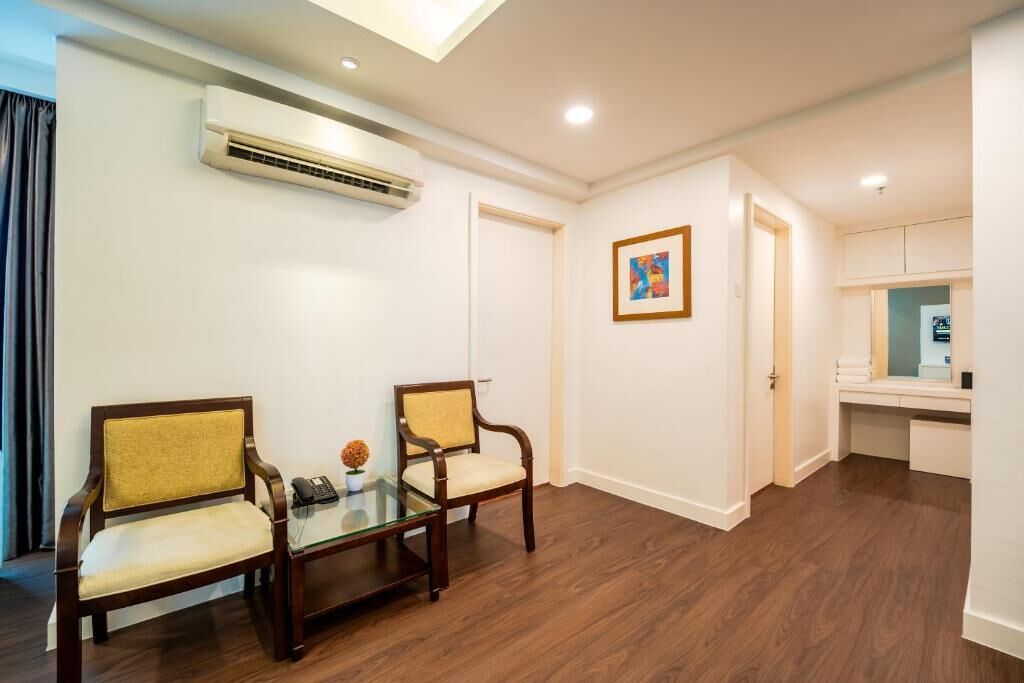 Quarto de hotel com piso de madeira, mesa de vidro, duas cadeiras, unidade de ar condicionado, quadro na parede e banheiro com espelho e porta-toalhas.