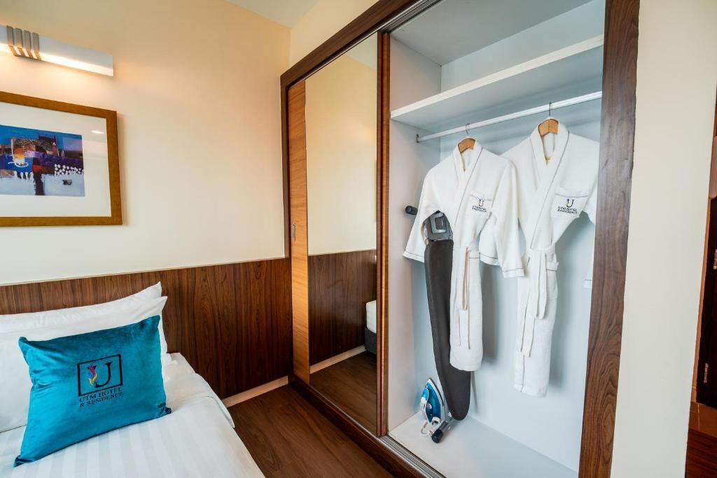 Quarto de hotel com uma cama, um travesseiro azul, um espelho e um guarda-roupa com roupões brancos.