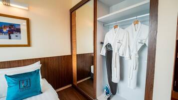 Quarto de hotel com uma cama, um travesseiro azul, um espelho e um guarda-roupa com roupões brancos.