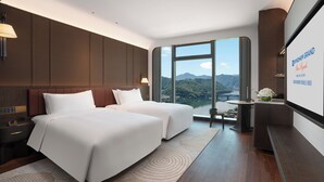 Quarto de hotel com duas camas, uma janela grande com vista para as montanhas, uma mesinha com um vaso de flores e uma televisão na parede.