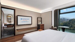 Quarto de hotel moderno com uma cama grande, uma escrivaninha, uma cadeira, uma mesinha, um espelho e uma televisão exibindo informações do hotel.