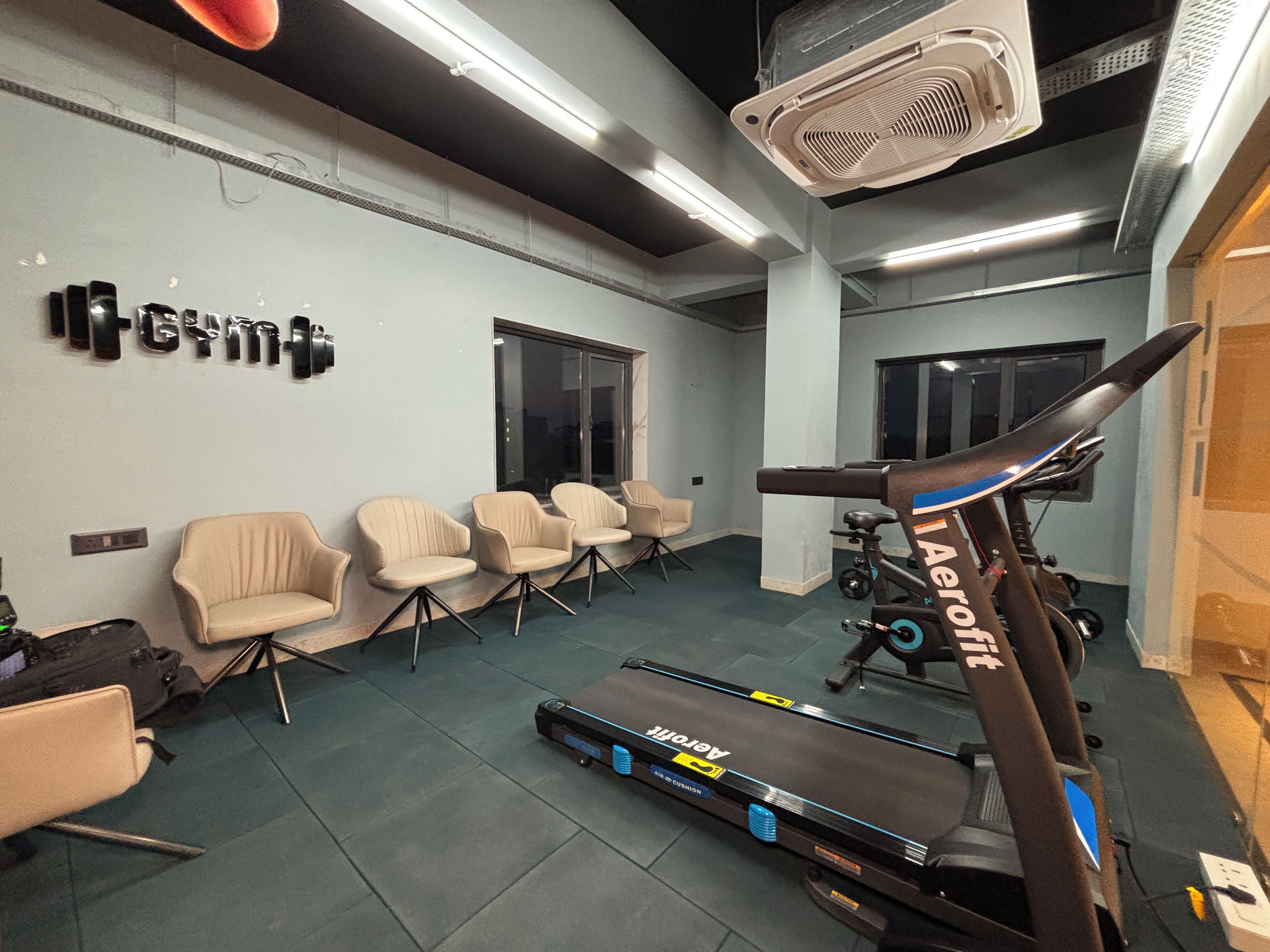 Academia com equipamentos de ginástica, incluindo esteira e bicicletas ergométricas, e algumas cadeiras.