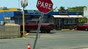 Vista da rua com uma placa de pare vermelha, fachada azul de um estabelecimento e uma caminhonete vermelha.