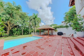 Uma piscina com um gazebo de telhado de palha, cercada por vegetação exuberante e um edifício branco.