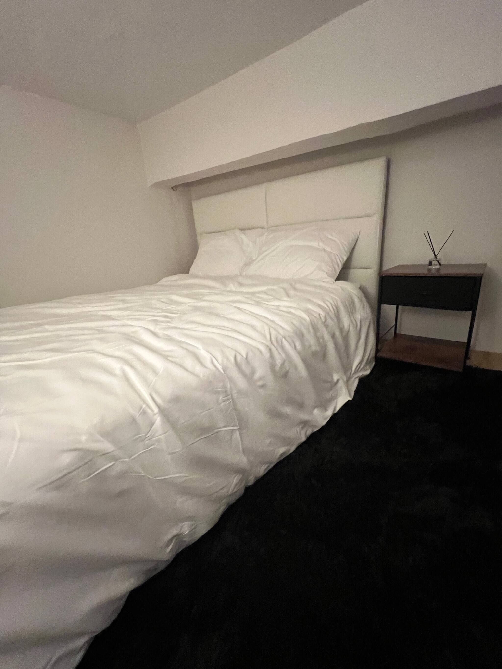 Uma cama de solteiro com roupa de cama branca num quarto com chão escuro.