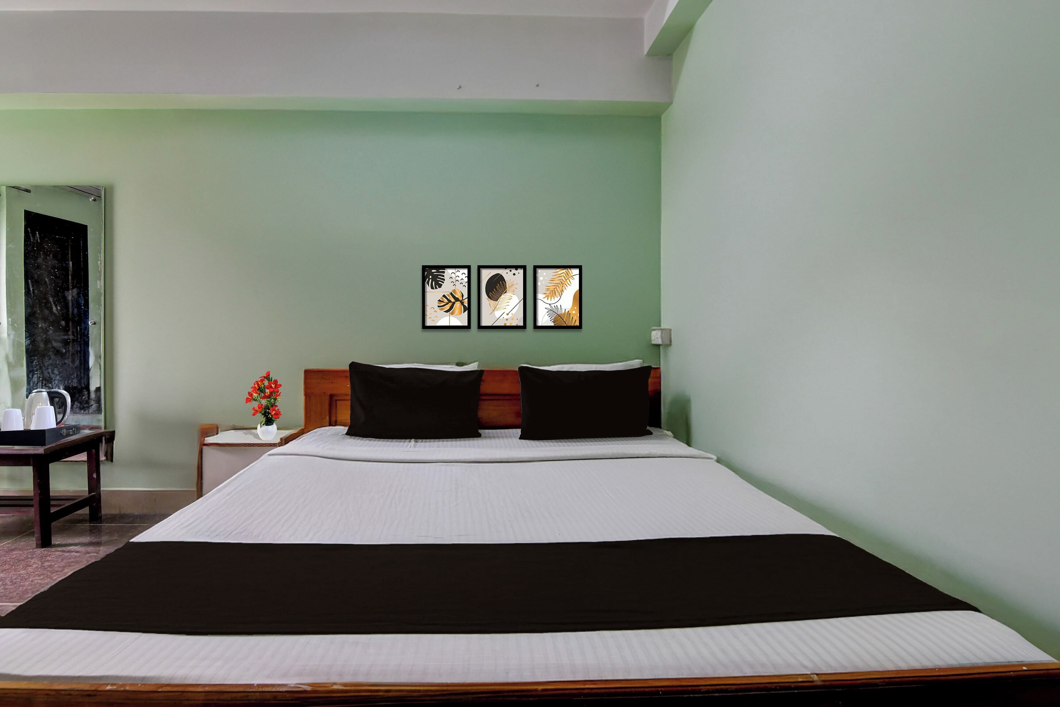 Quarto de hotel com uma cama grande, duas almofadas pretas, uma mesa de cabeceira com um vaso de flores e duas obras de arte emolduradas na parede.