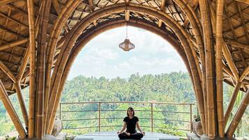 Uma pessoa meditando em um quarto com teto de bambu e vista para uma paisagem exuberante.