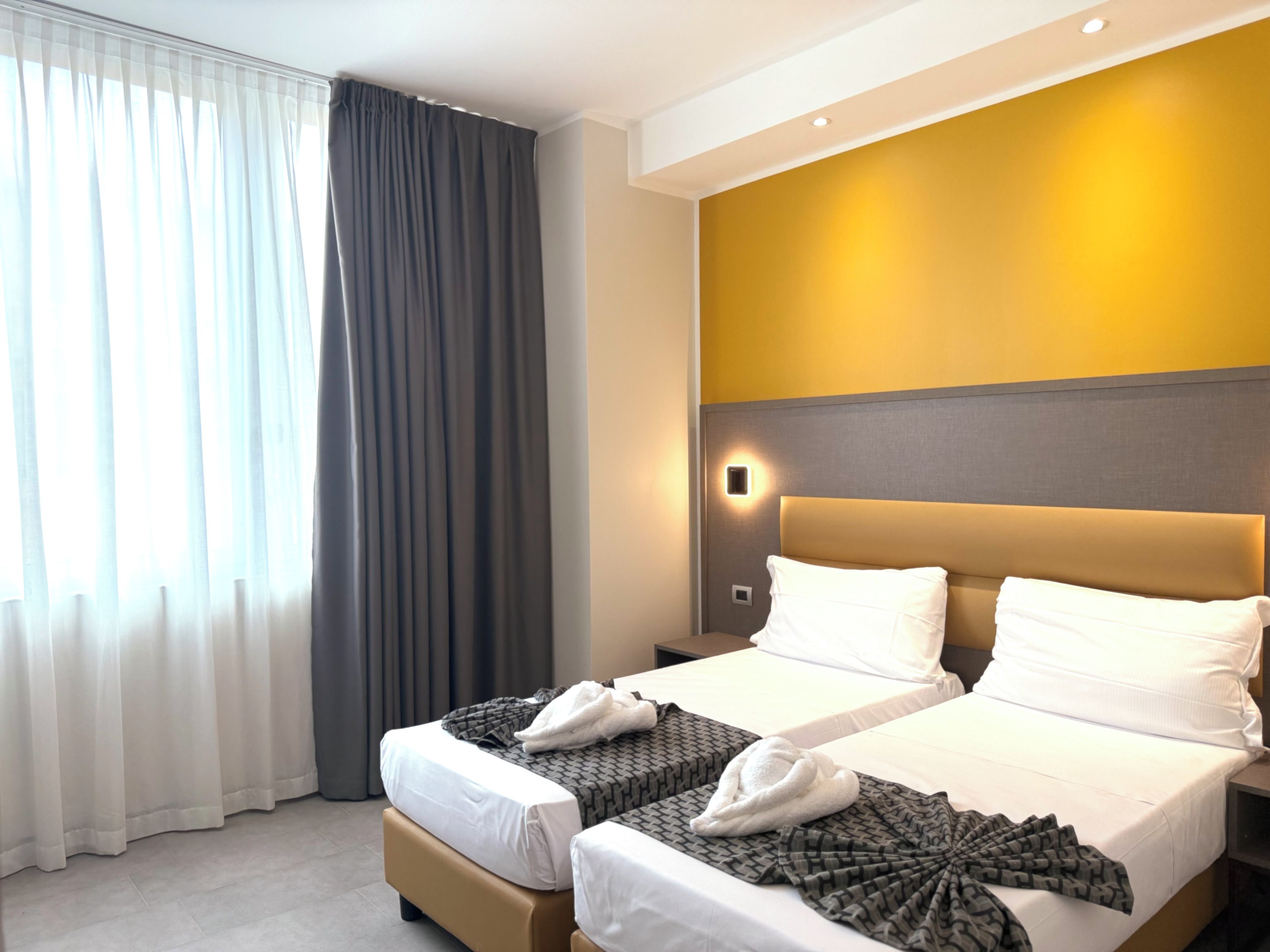 Quarto de hotel com duas camas, uma janela grande com cortinas, uma luminária fixada na parede e uma parede lisa com detalhes em amarelo.